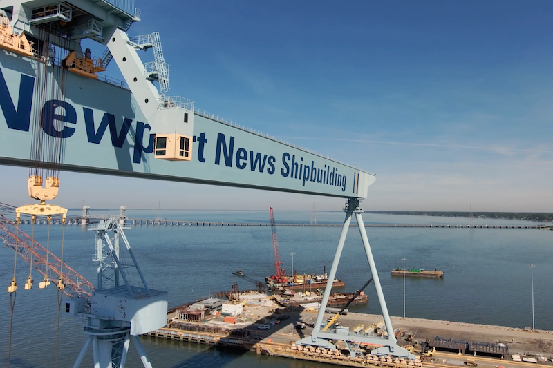 The image shows a large gantry crane at the Newport News Shipbuilding facility. The crane is ...