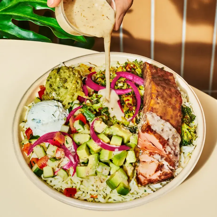 Hand pouring dressing over a CAVA Grains Bowl with salmon, pickled ...