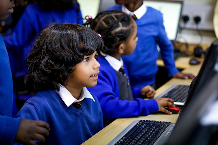 Primary school child in uniform sits at a desk in front of a silver laptop.